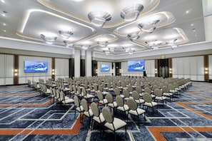 Sala de conferências com fileiras de cadeiras, grandes telas e iluminação moderna no teto.