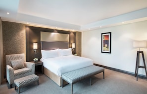 Quarto de hotel com cama, cadeira, banqueta, mesa de cabeceira com abajur e um quadro na parede.