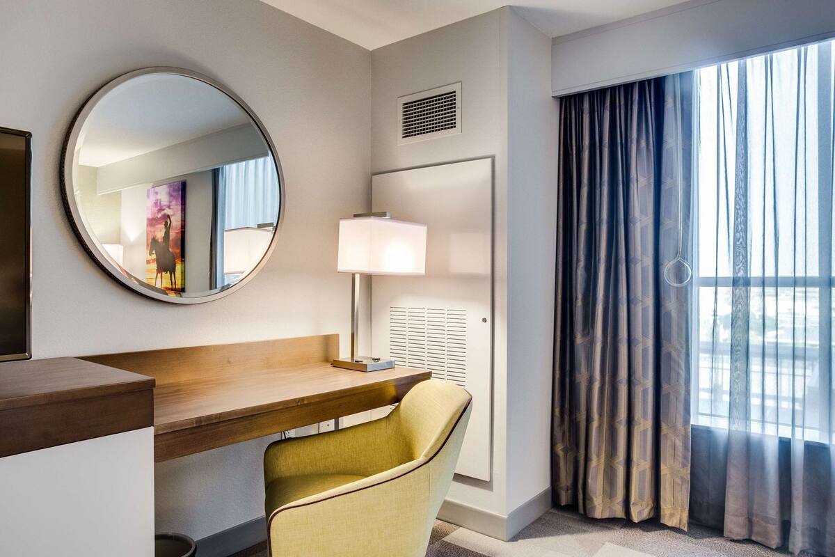 Quarto de hotel com escrivaninha, cadeira, espelho redondo, abajur e uma janela grande com cortinas.