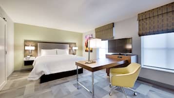 Quarto de hotel com cama, escrivaninha, cadeira, televisão e janela com persianas.