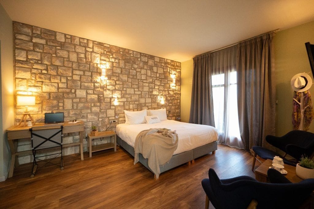 Quarto com parede de pedra, cama, escrivaninha com laptop, cadeira e janela com cortinas.