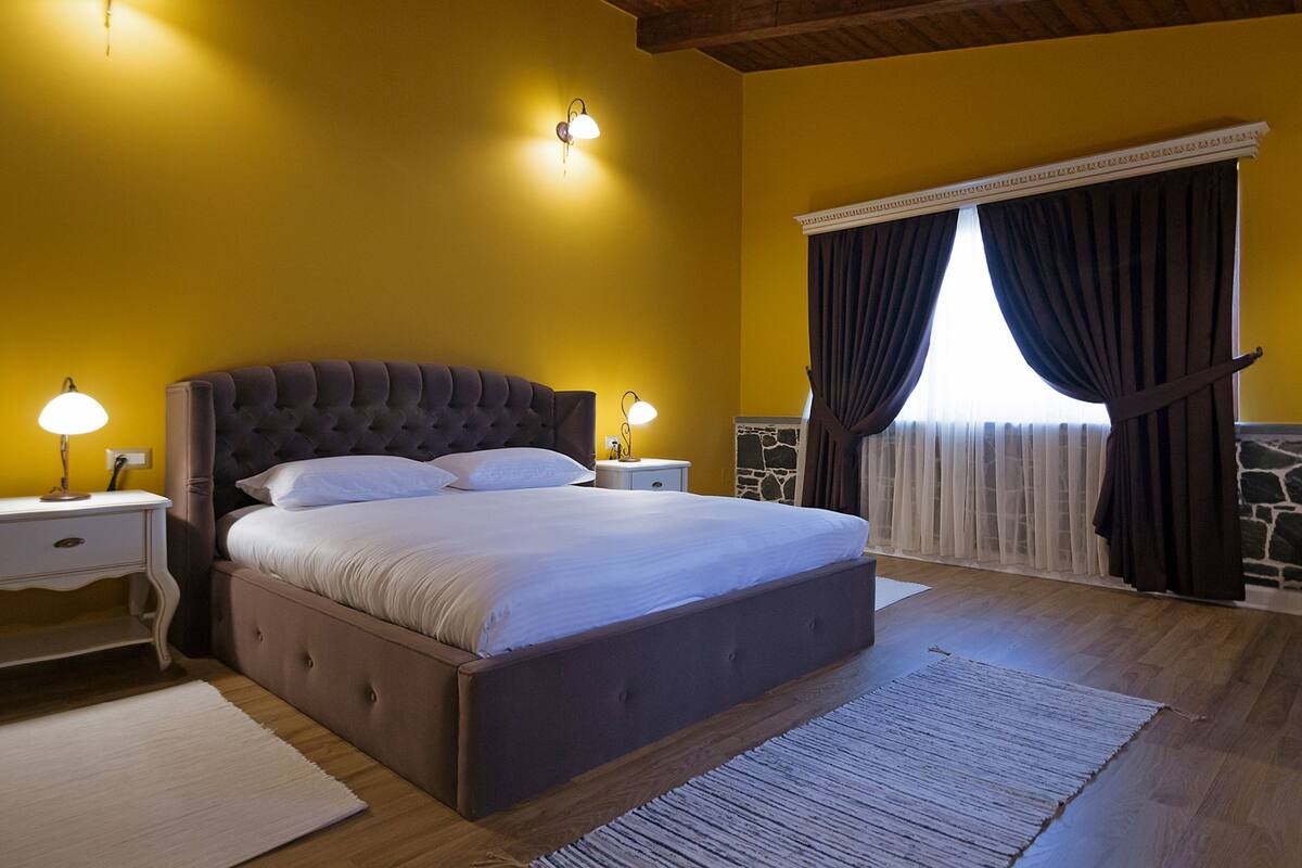 Quarto com uma cama grande, duas luminárias de cabeceira, uma mesa de cabeceira, uma janela com cortinas e piso de madeira.