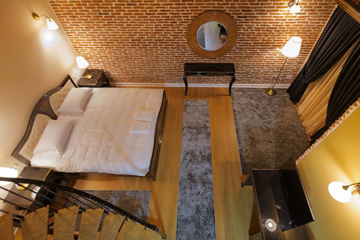 Quarto de hotel com parede de tijolos, cama com lençóis brancos, uma pequena mesa, um espelho e uma escada.