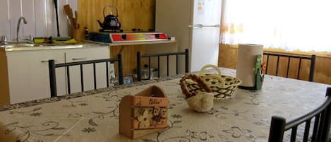 Uma cozinha compacta com micro-ondas, frigorífico e uma pia pequena. Há uma mesa com toalha florida e uma cesta de pão.
