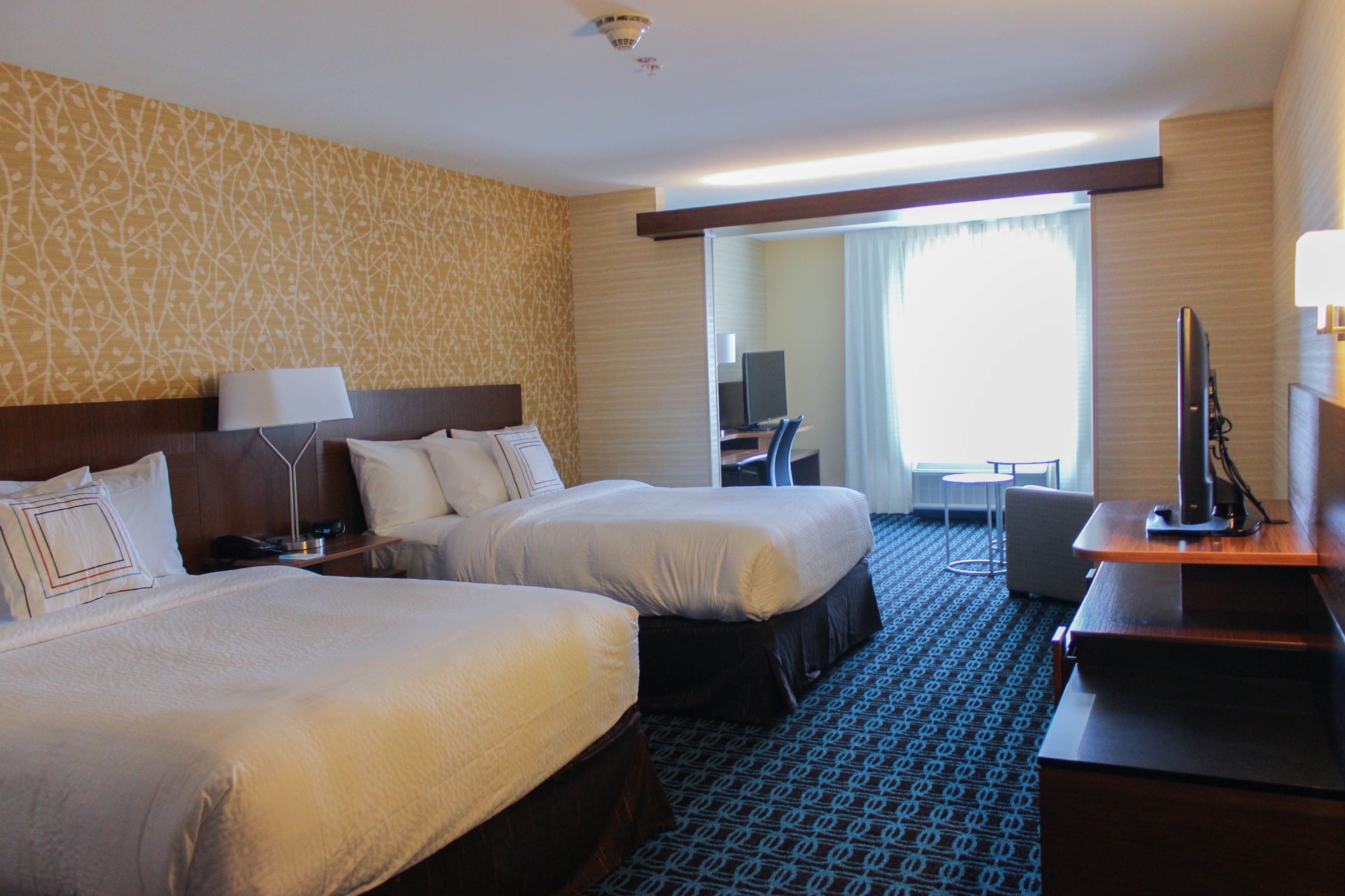 Quarto de hotel com duas camas, uma escrivaninha, uma cadeira, uma TV e uma pequena mesa.