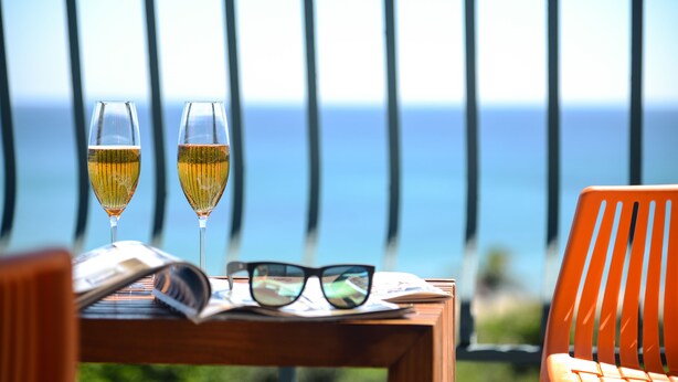 Dois copos de vinho branco sobre uma mesa de madeira, com óculos de sol e uma revista, com vista para a paisagem oceânica.