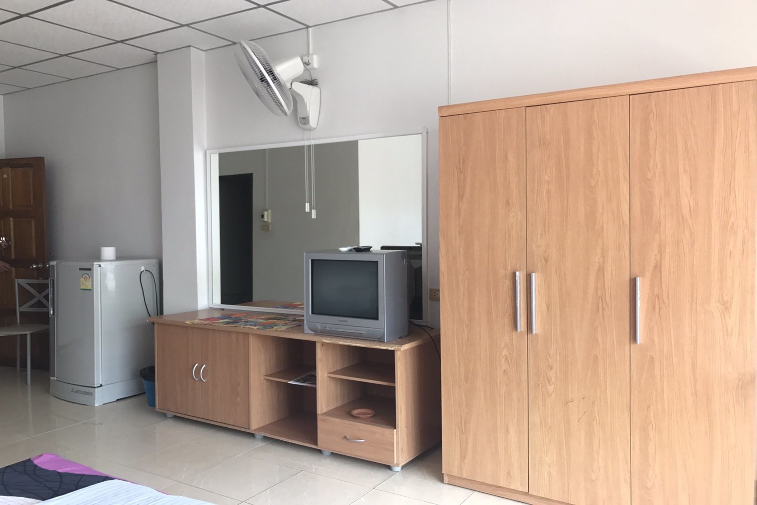 Quarto com armário de madeira, televisão em um suporte, geladeira e espelho.
