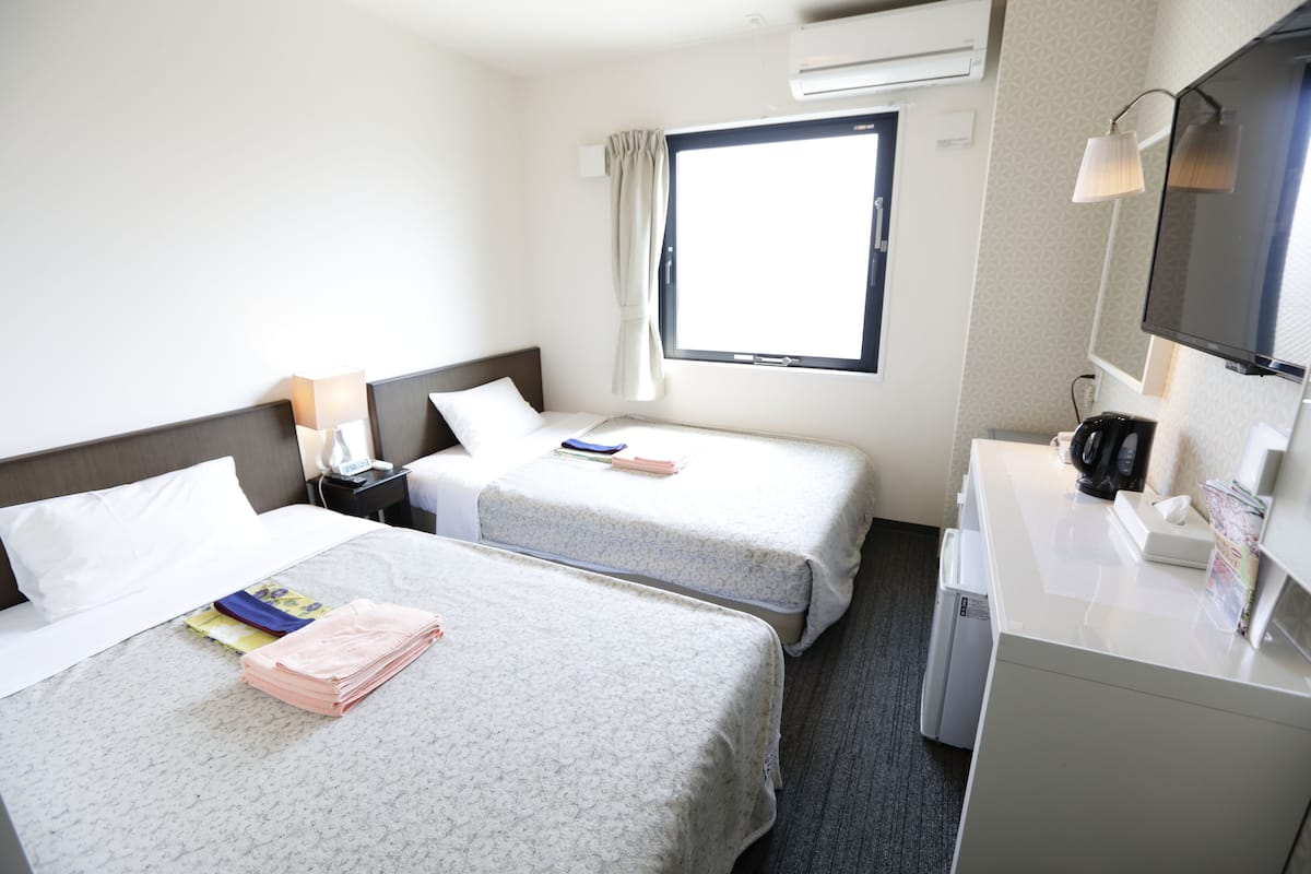 Quarto de hotel com duas camas, uma escrivaninha, um espelho, uma janela com cortinas e um ar condicionado.