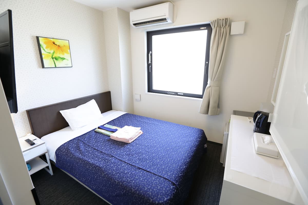 Quarto de hotel com cama, mesa de cabeceira, escrivaninha, janela com cortinas e ar condicionado.