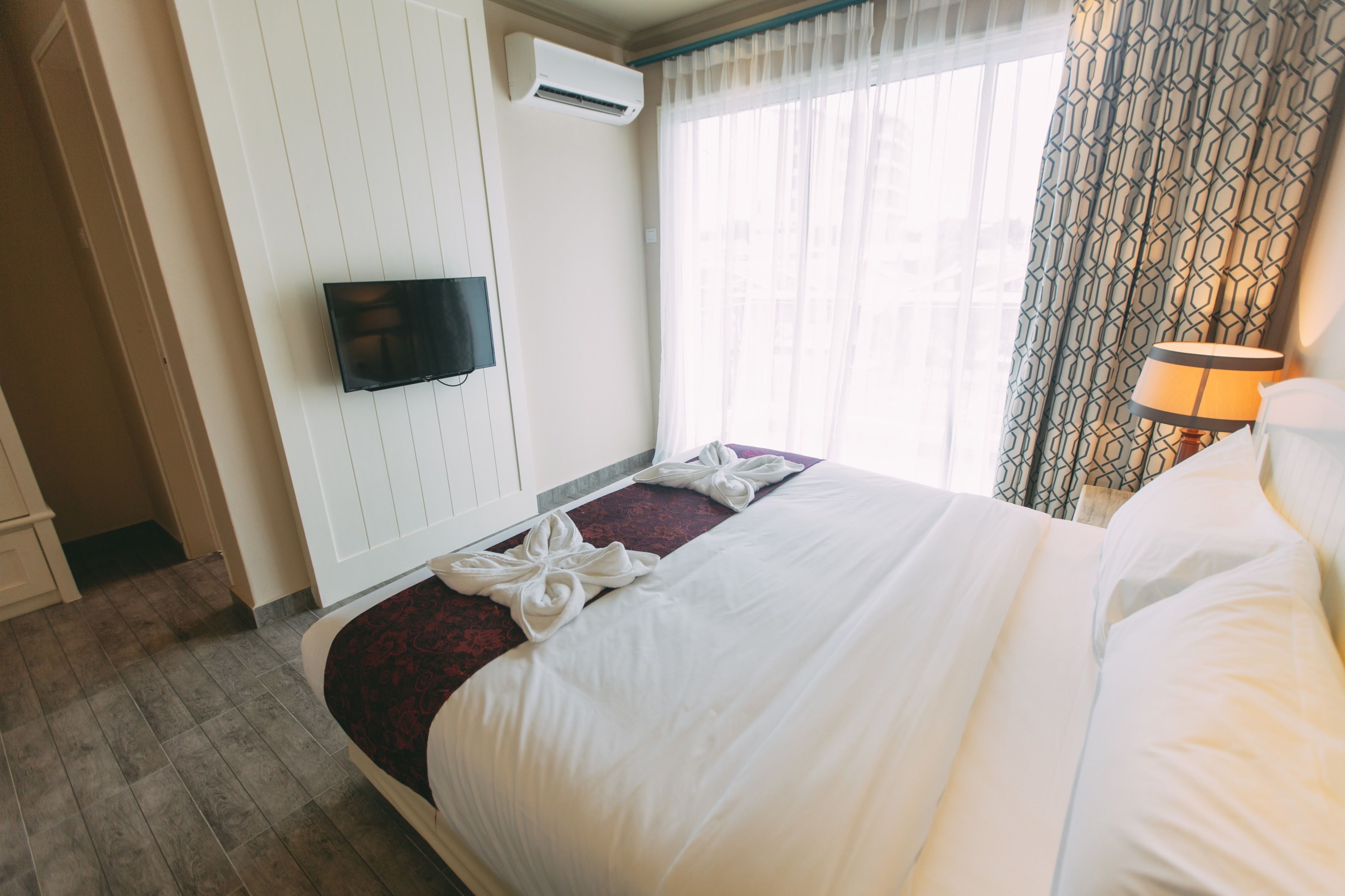 Quarto de hotel com cama, televisão de tela plana na parede, aparelho de ar condicionado e janela com cortinas leves.
