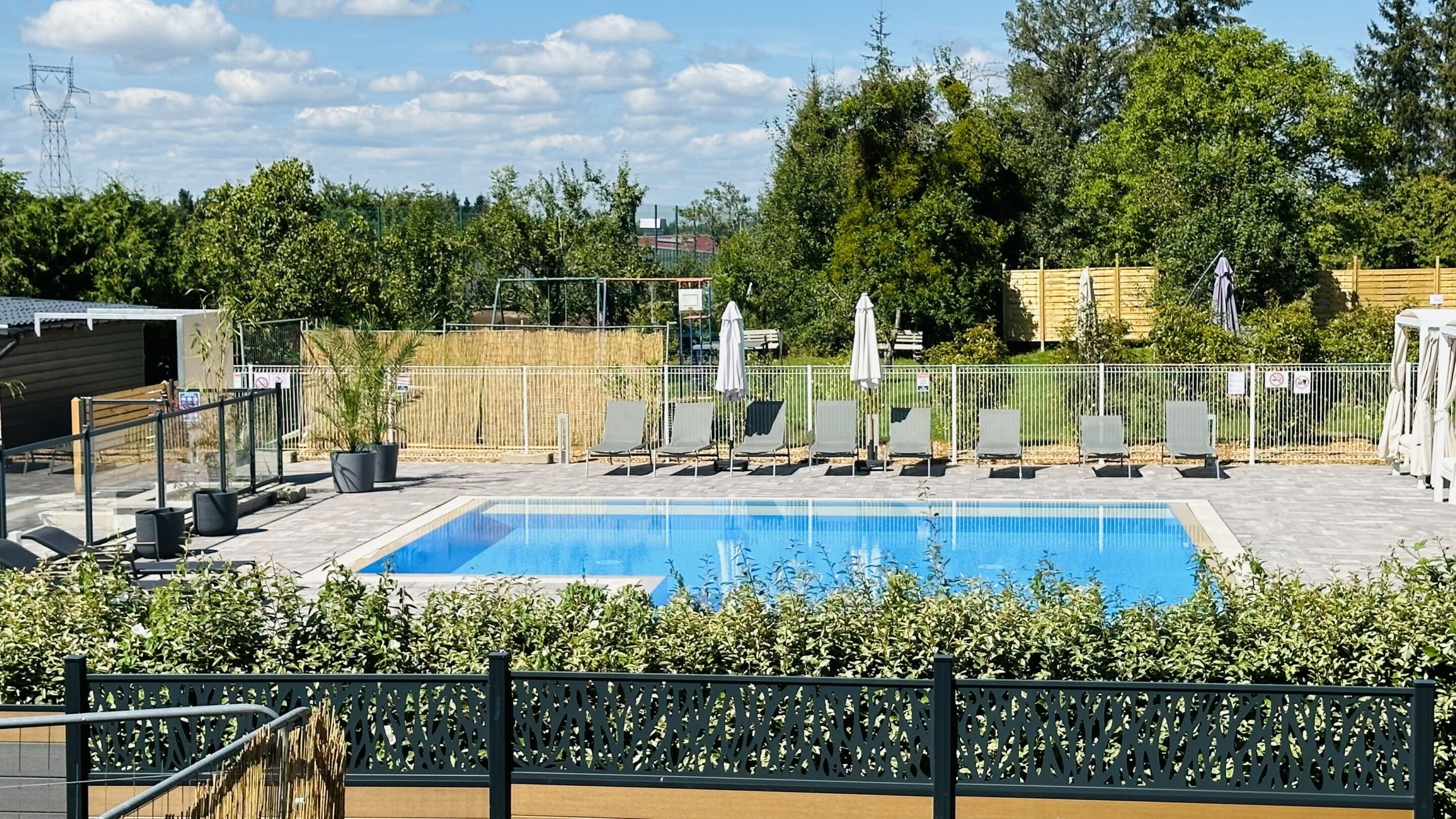 Piscina retangular com espreguiçadeiras e guarda-sóis, rodeada por uma cerca e vegetação.