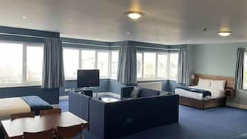 Quarto de hotel com cama, sofá, escrivaninha com cadeiras, televisão e grandes janelas.