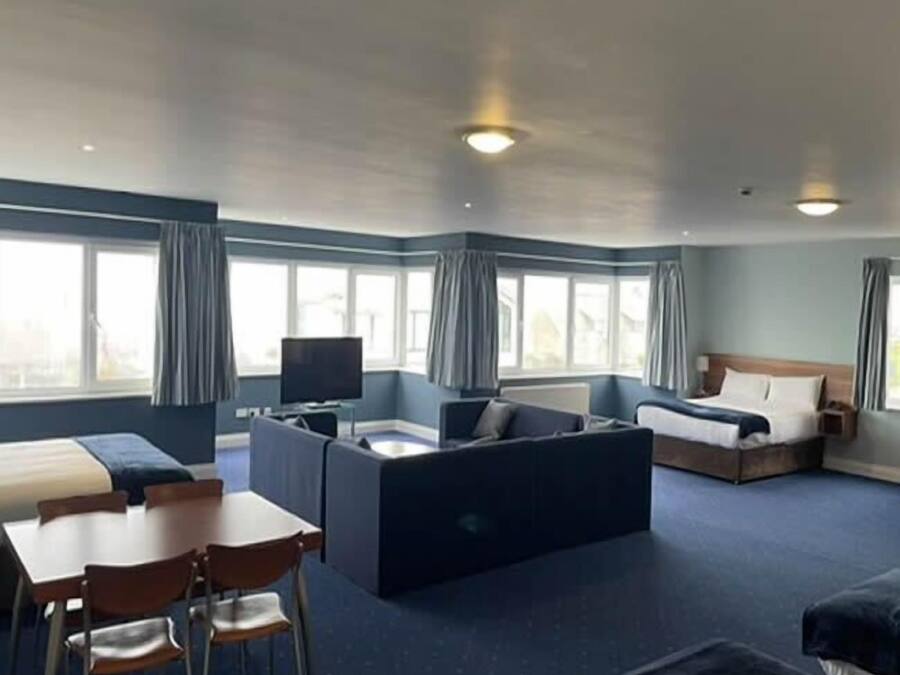 Quarto de hotel com cama, sofá, escrivaninha com cadeiras, televisão e grandes janelas.