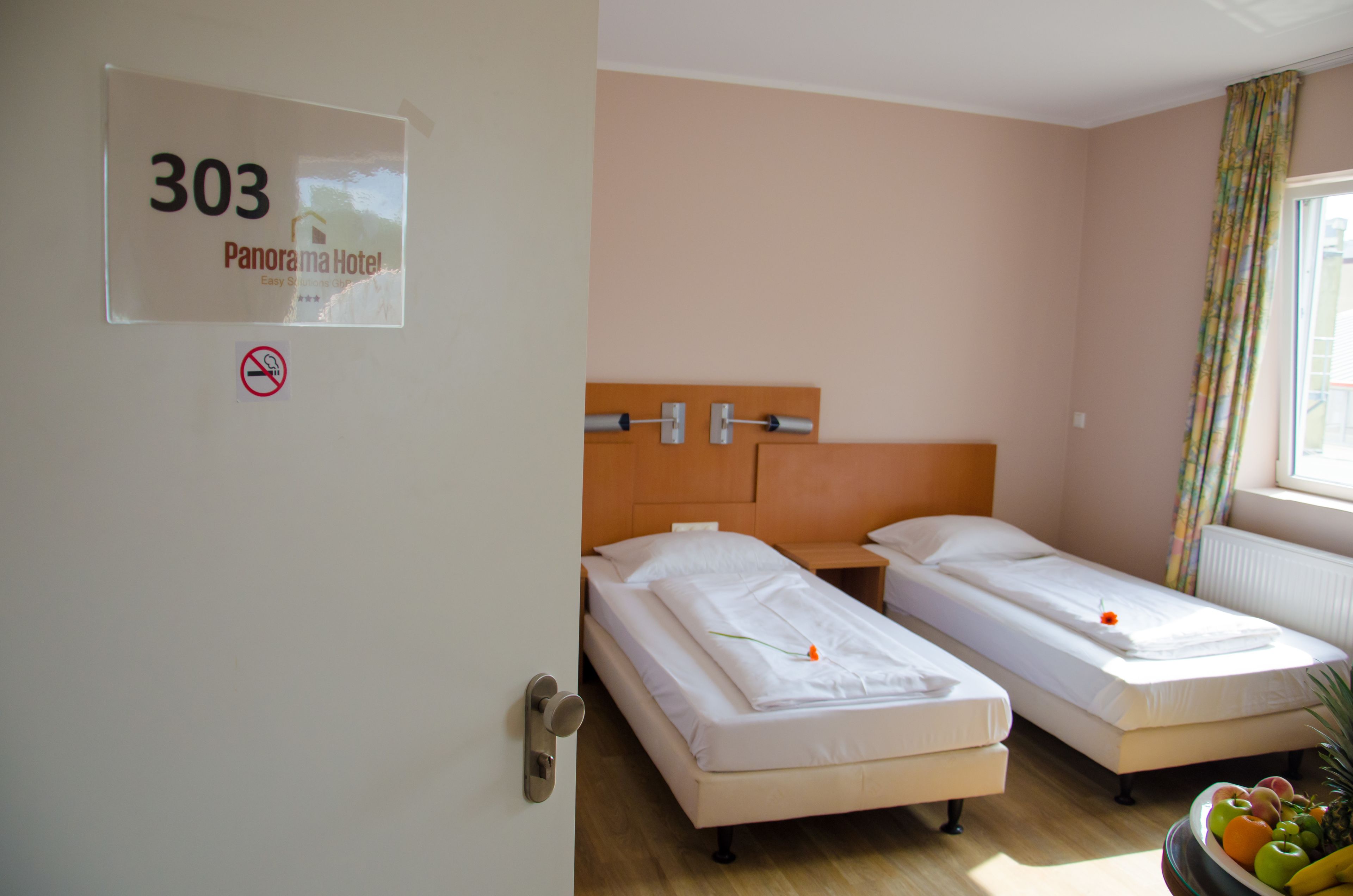 Quarto de hotel com duas camas de solteiro, cabeceira de madeira, mesa de cabeceira pequena, tigela de frutas à direita e uma placa indicando o número do quarto 303.