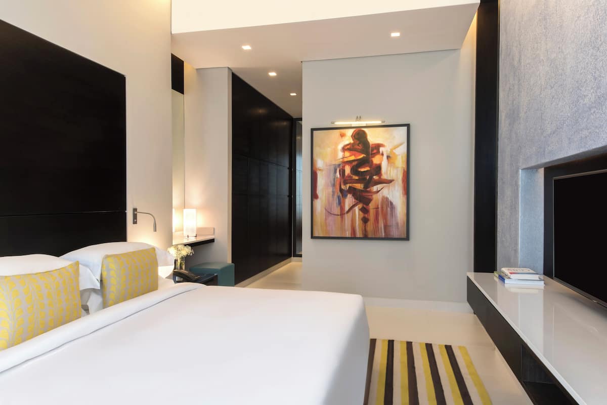 Quarto de hotel moderno com uma cama grande, televisão de tela plana, um quadro na parede e uma mesinha de cabeceira com abajur.