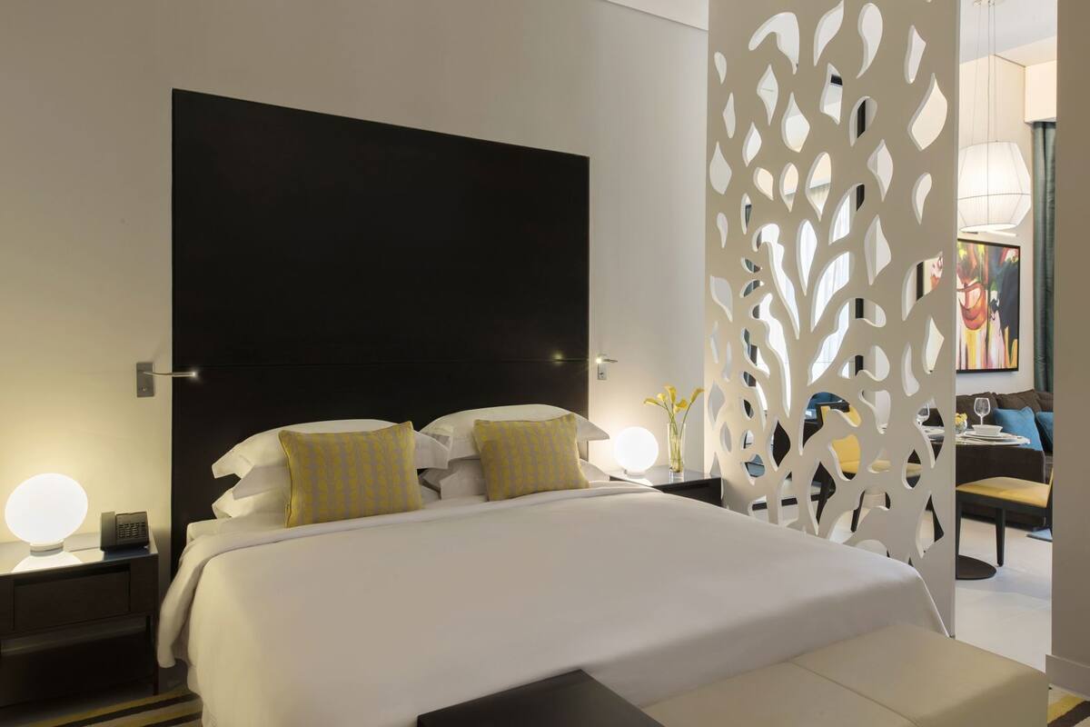 Quarto de hotel moderno com uma cama grande, mesas de cabeceira, um divisória branca decorativa e vista para a sala de estar.