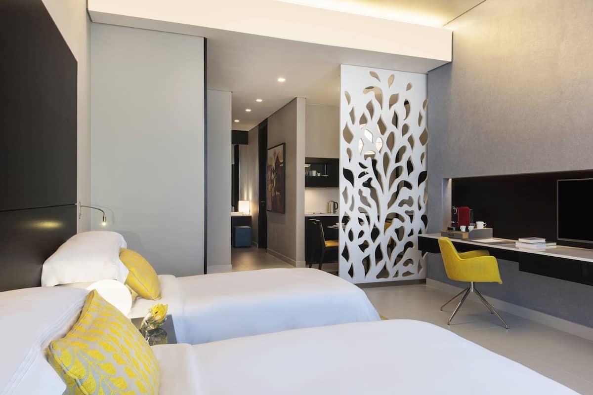 Quarto de hotel moderno com duas camas, uma escrivaninha, uma cadeira, uma TV e um separador decorativo.