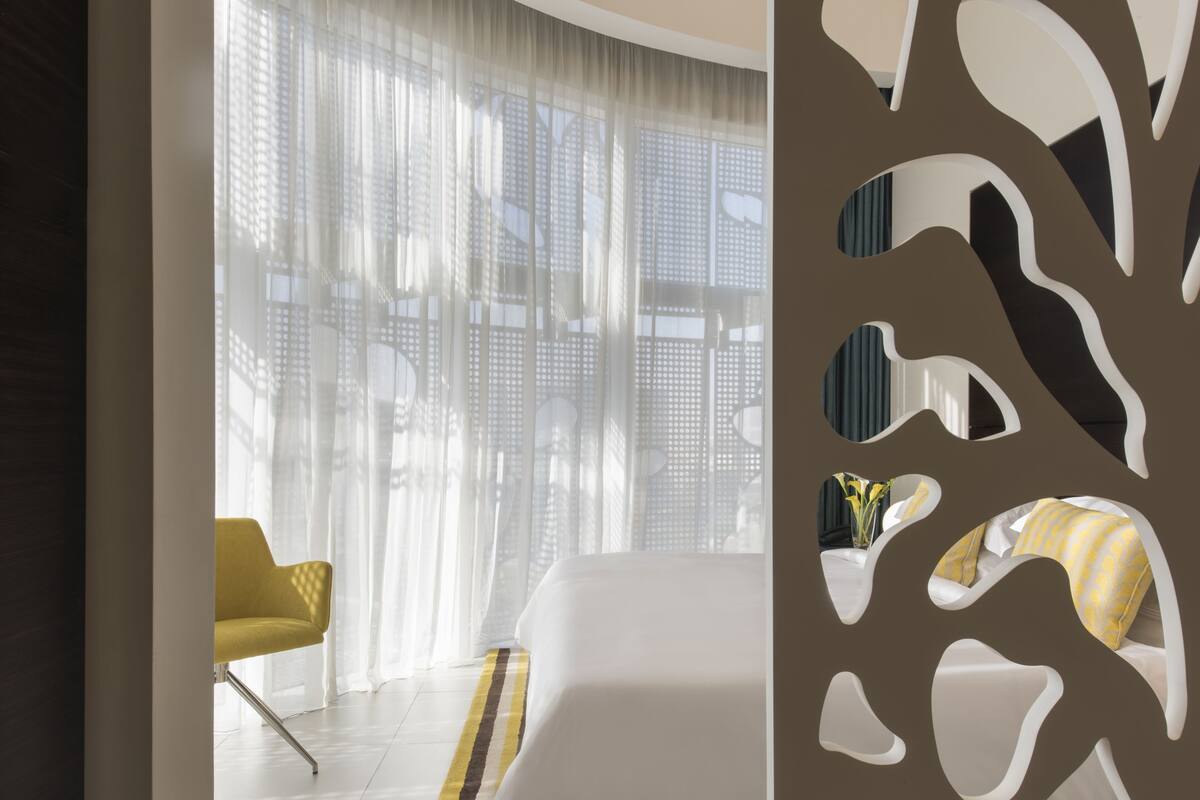 Quarto de hotel moderno com uma cama grande, uma poltrona amarela e cortinas leves que permitem a entrada de luz natural.
