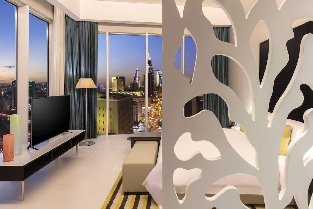 Quarto de hotel moderno com uma televisão grande, um sofá, uma mesinha e vista para o horizonte da cidade ao entardecer.