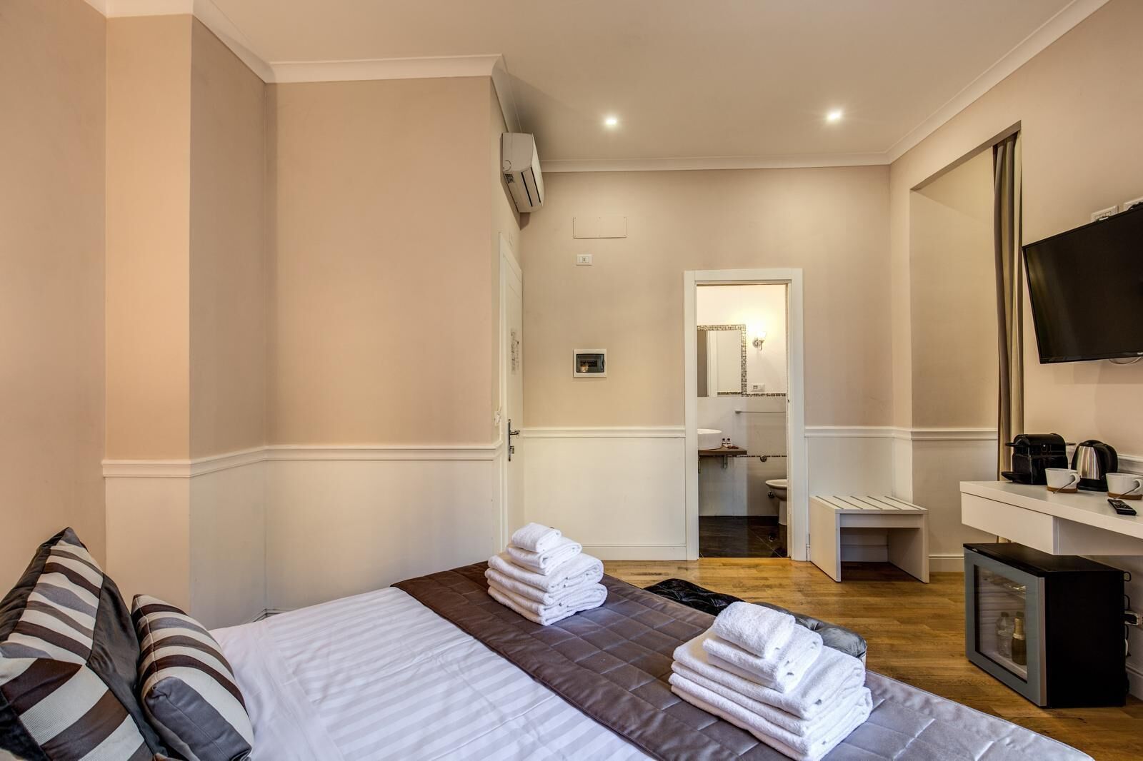 Quarto de hotel com cama, televisão, mesa com cafeteira e banheiro visível por uma porta aberta.