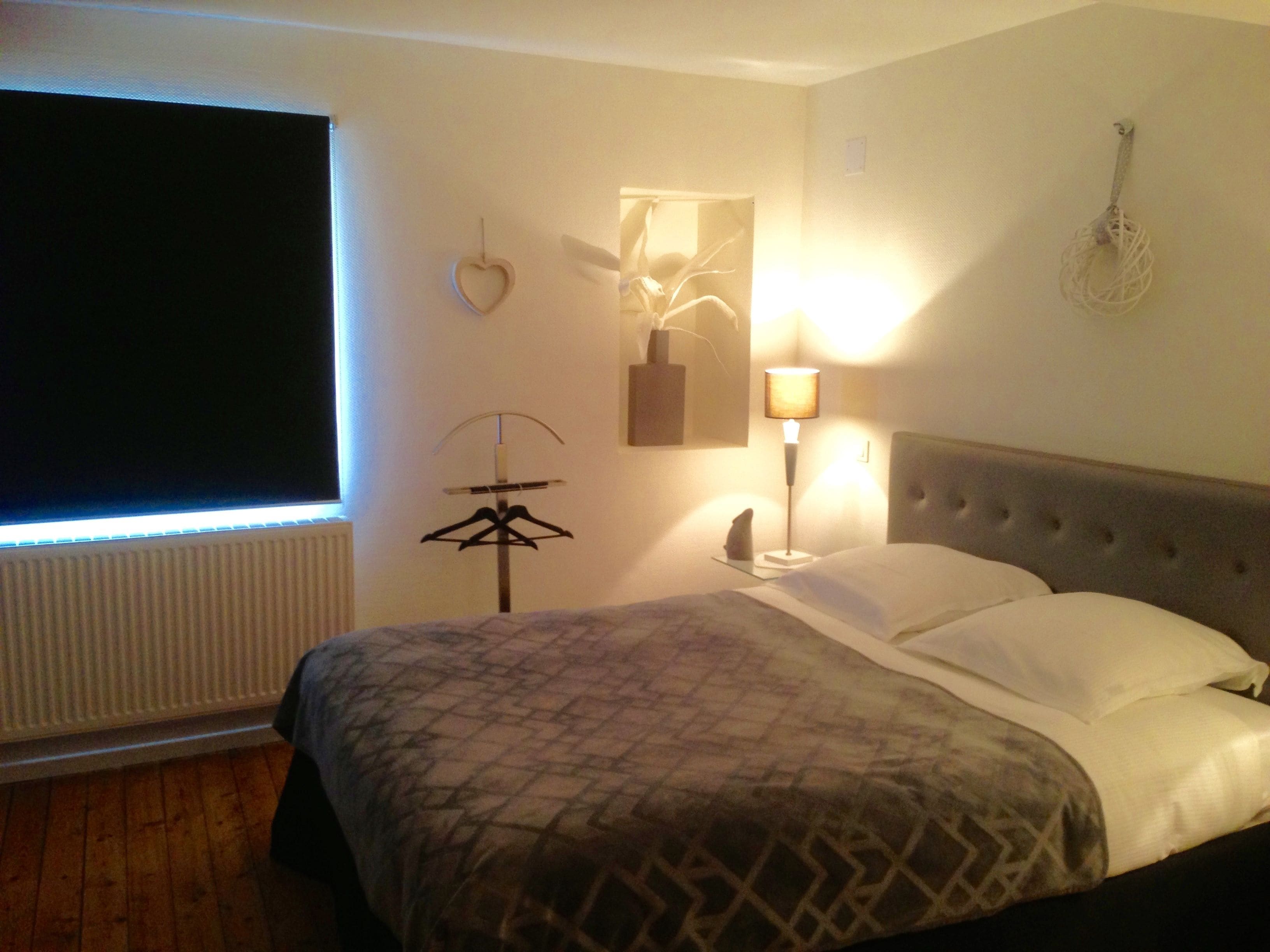 Quarto bem arrumado com uma cama, abajures, radiador, decoração de coração na parede e uma janela com persianas.
