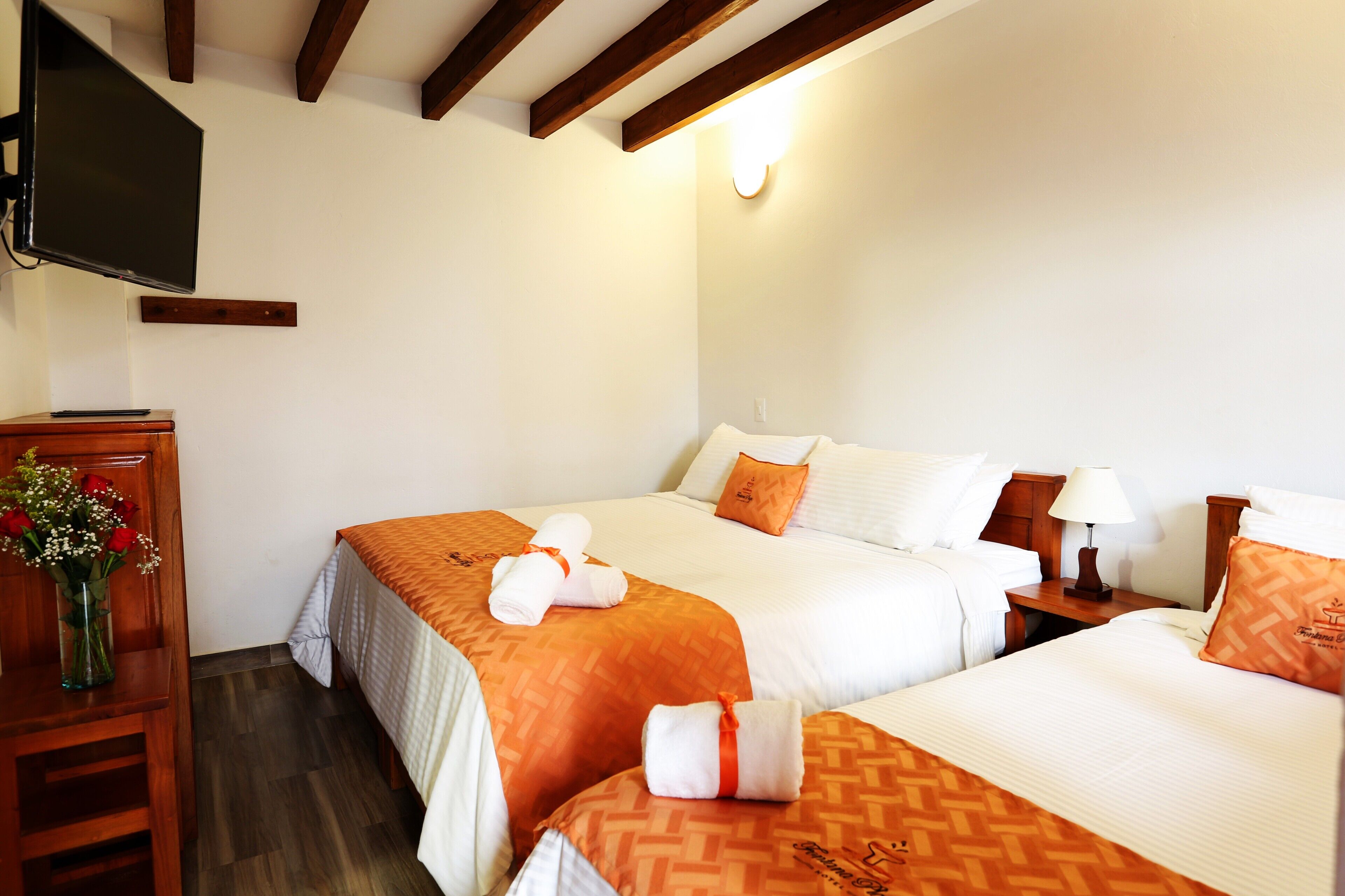 Quarto de hotel com duas camas, TV fixada na parede, escrivaninha de madeira e um vaso com flores.