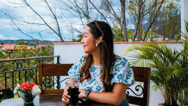 Mulher sentada à mesa de madeira com uma bebida, ao ar livre, com vista para árvores e edifícios.