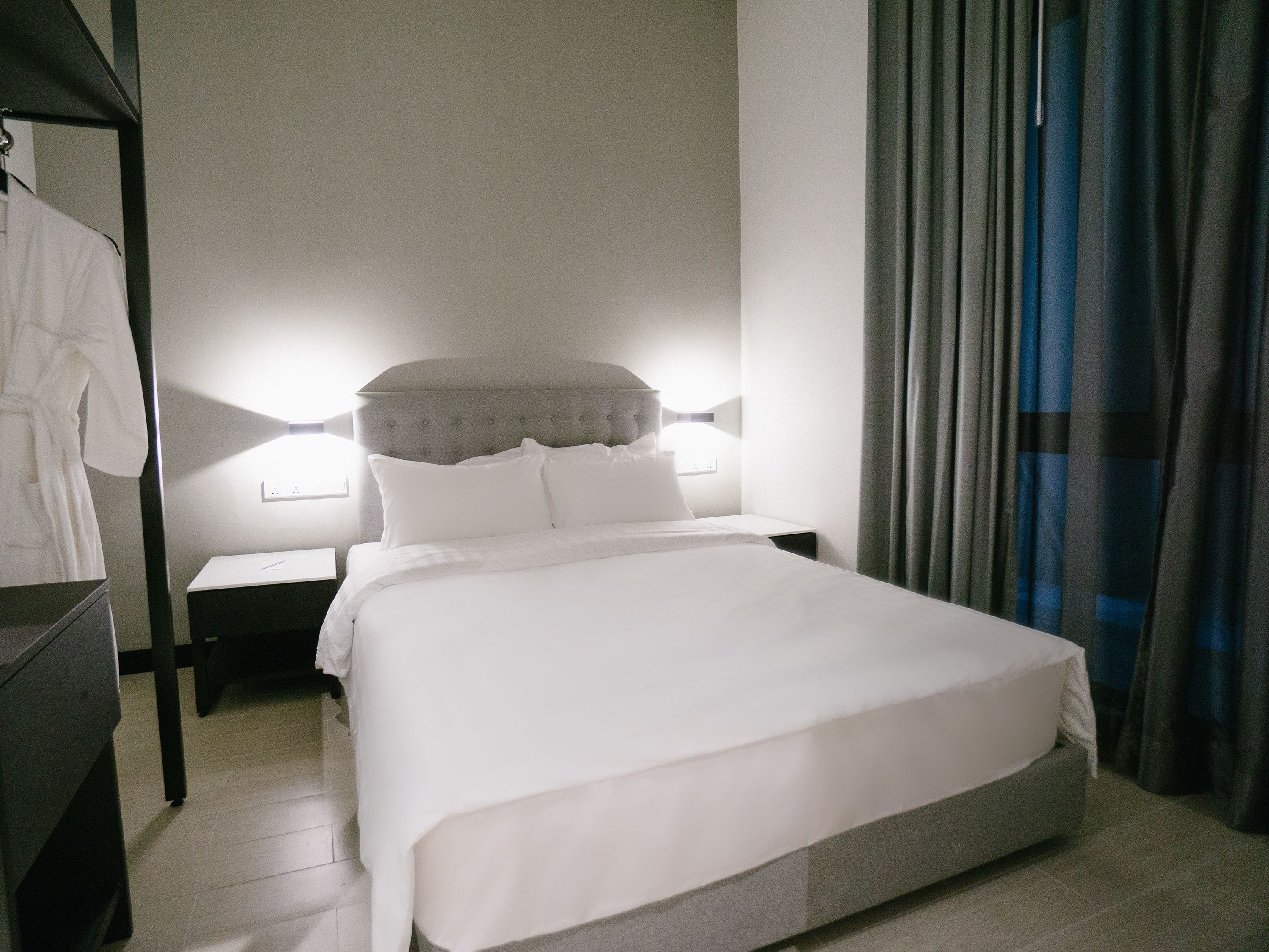 Quarto de hotel com uma cama grande, uma mesa de cabeceira, um roupão pendurado em um suporte e uma janela com cortinas.