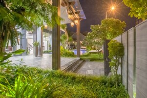 Uma área externa com piscina, plantas em vasos e um caminho que leva à entrada de um edifício.