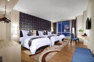 Quarto de hotel com duas camas, uma janela grande, uma escrivaninha com cadeira e televisão fixada na parede.