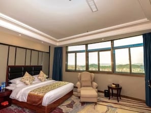 Quarto de hotel com uma cama grande, uma cadeira, uma mesinha e vista para a paisagem urbana através de janelas amplas.