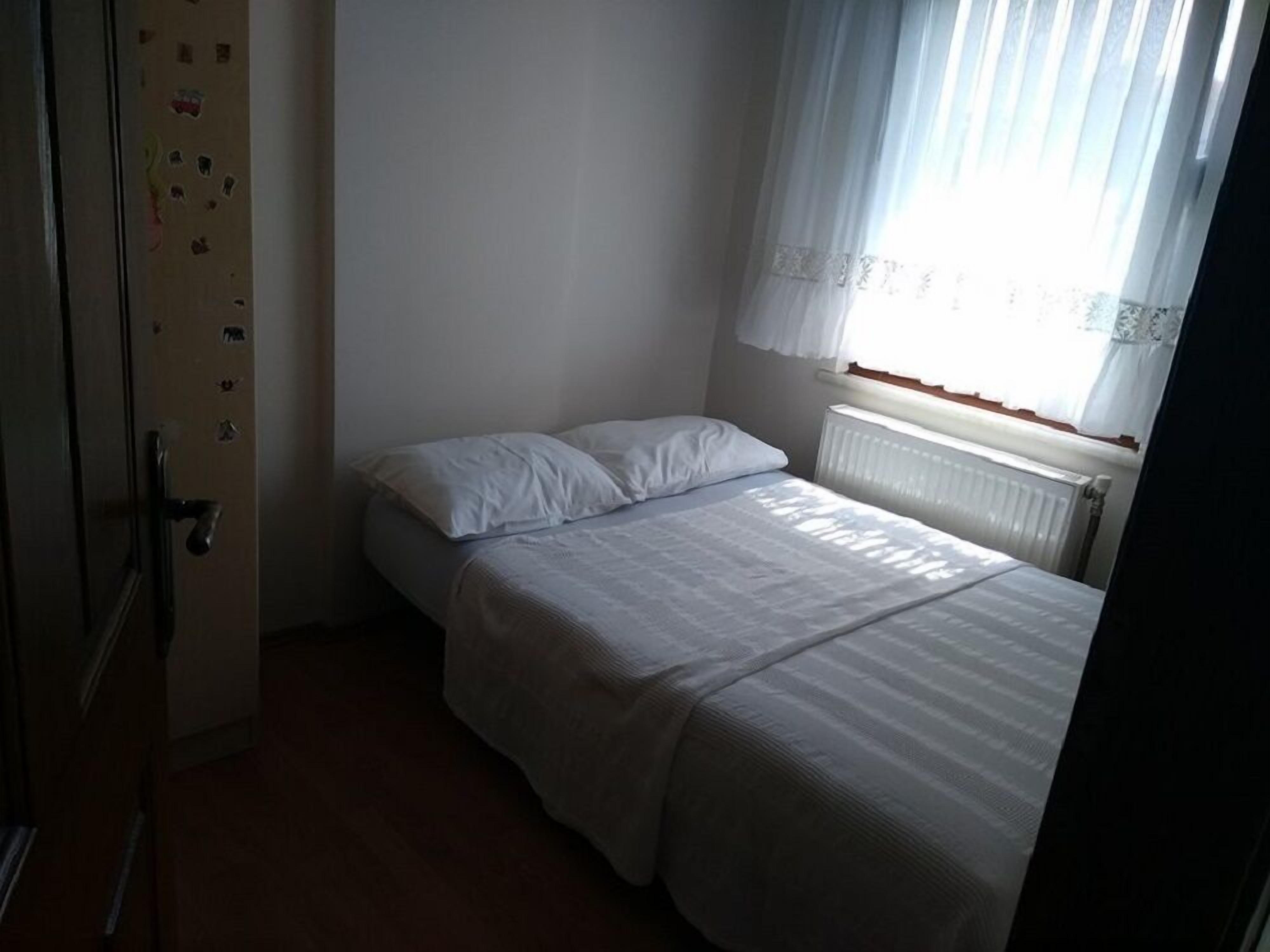 Quarto pequeno e simples, com uma cama, um radiador e uma janela com cortinas leves.
