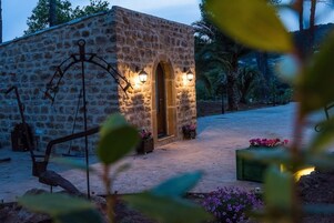 Um edifício de pedra com porta e duas luzes fixadas na parede, rodeado por um espaço pavimentado com flores e plantas.