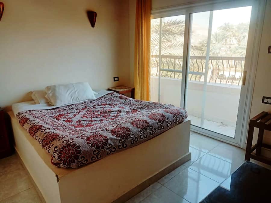 Quarto de hotel com cama, mesa de cabeceira, janela com vista para palmeiras e porta de correr para a varanda.