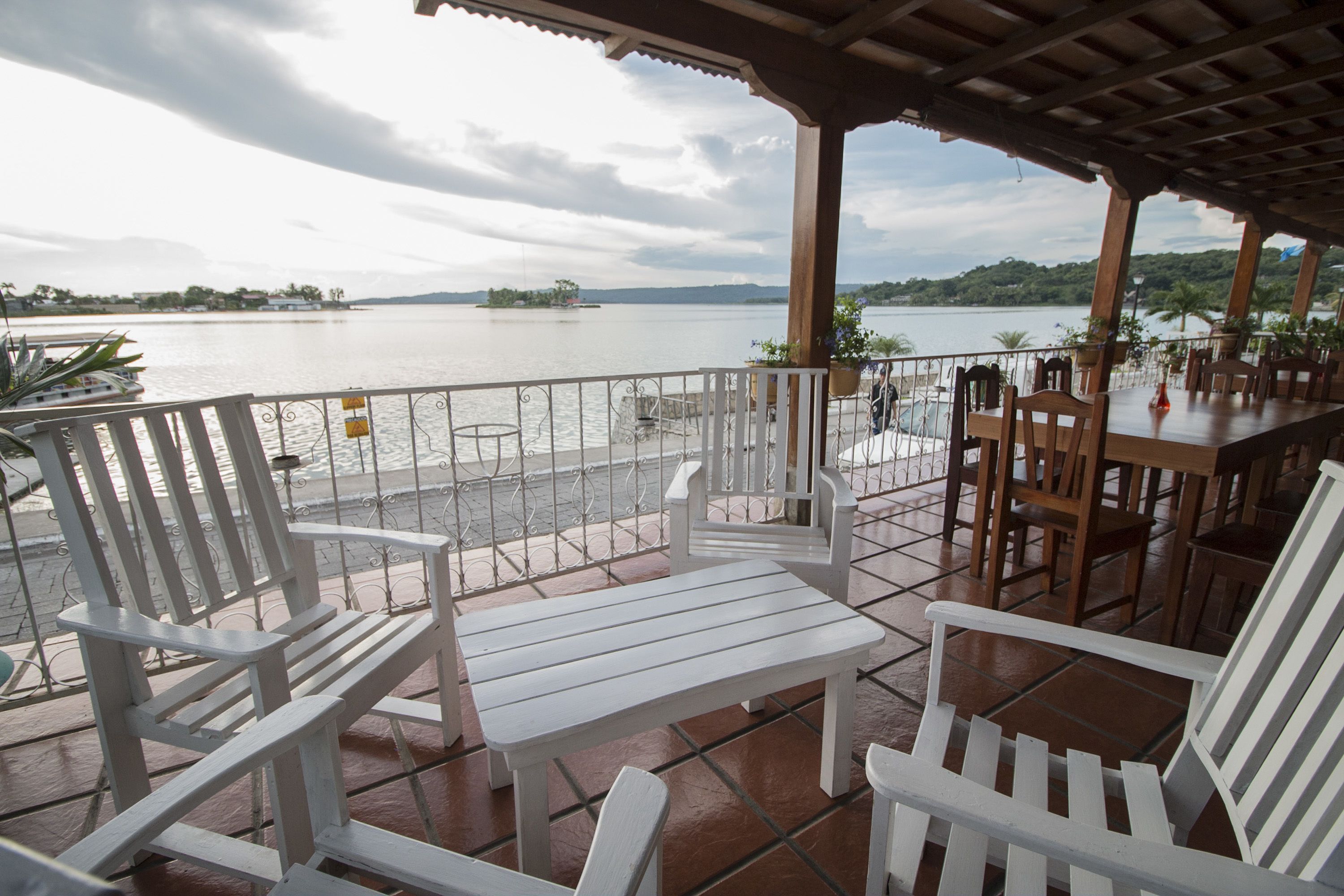Terraço com cadeiras e mesa brancas, com vista para um lago tranquilo com barcos e ilhas ao longe.
