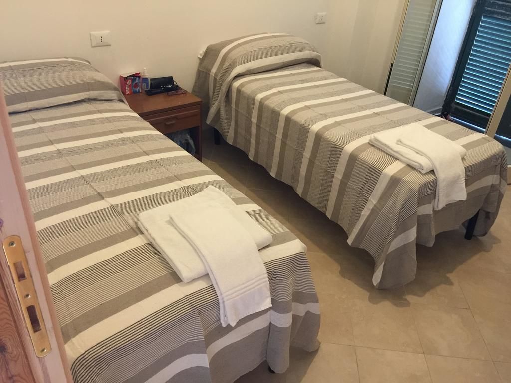 Quarto de hotel com duas camas de solteiro, uma mesinha de cabeceira de madeira com abajur e uma janela com persianas.