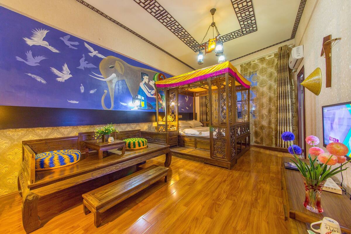 Quarto tradicional com chão de madeira, cama com dossel, um banco, uma pequena mesa com um vaso de flores e parede com um mural.