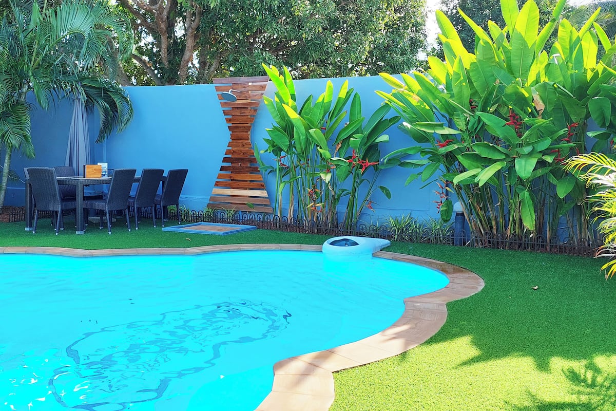 Piscina com deck de madeira e cadeiras, rodeada por vegetação exuberante e uma parede azul.