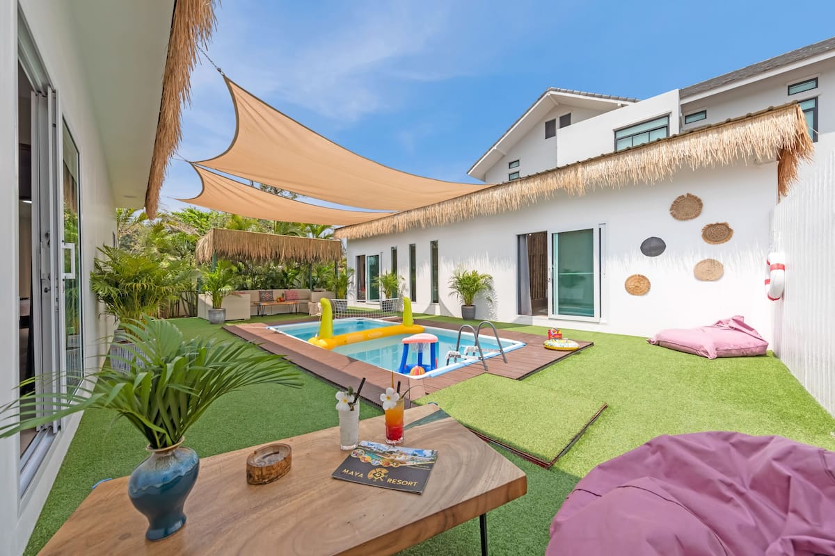 Um quintal com piscina, uma mesa de madeira com um livro e uma estrutura de telhado de palha.