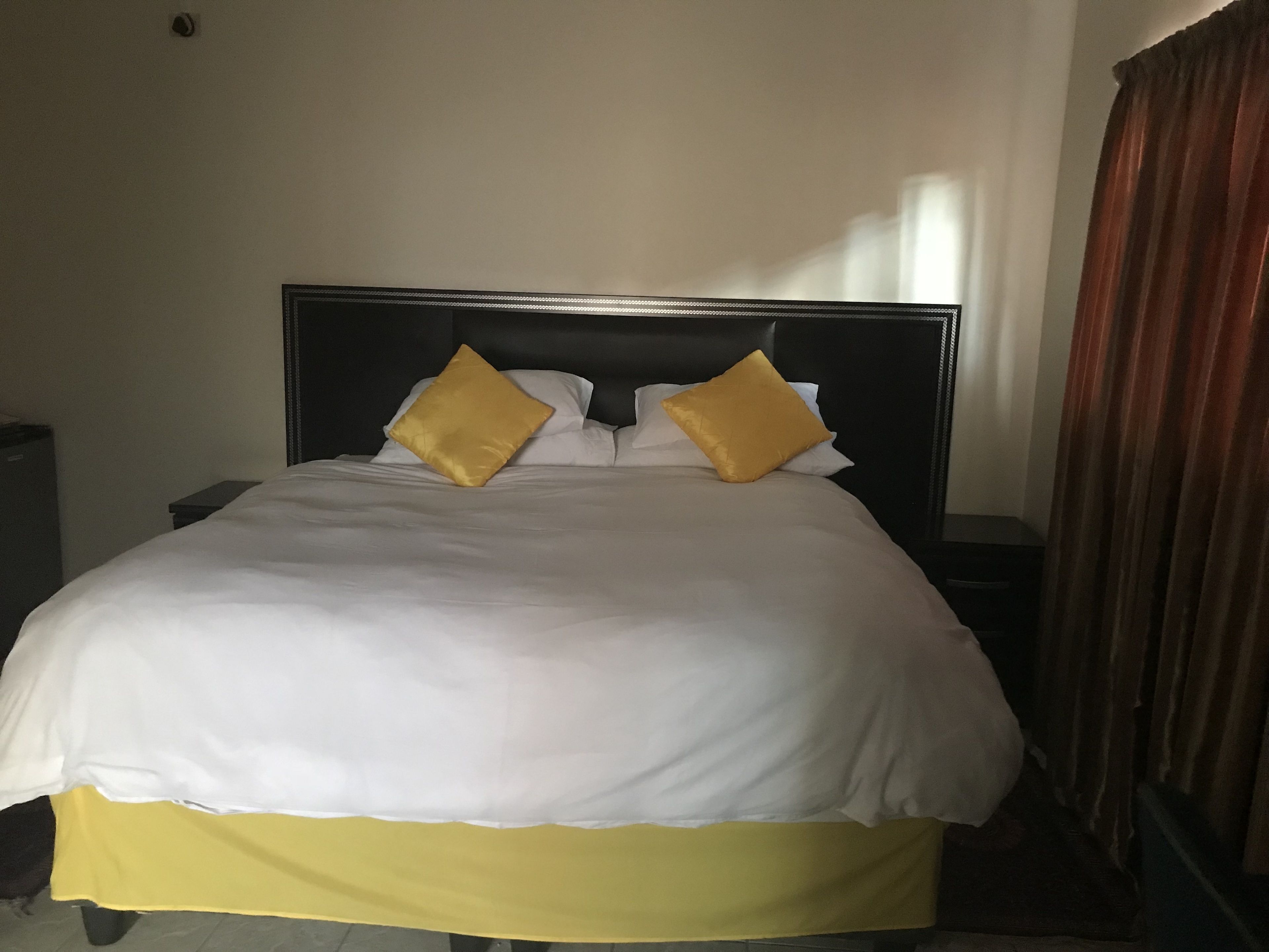 Cama com roupa de cama branca e duas almofadas amarelas, cabeceira preta e cortina marrom.