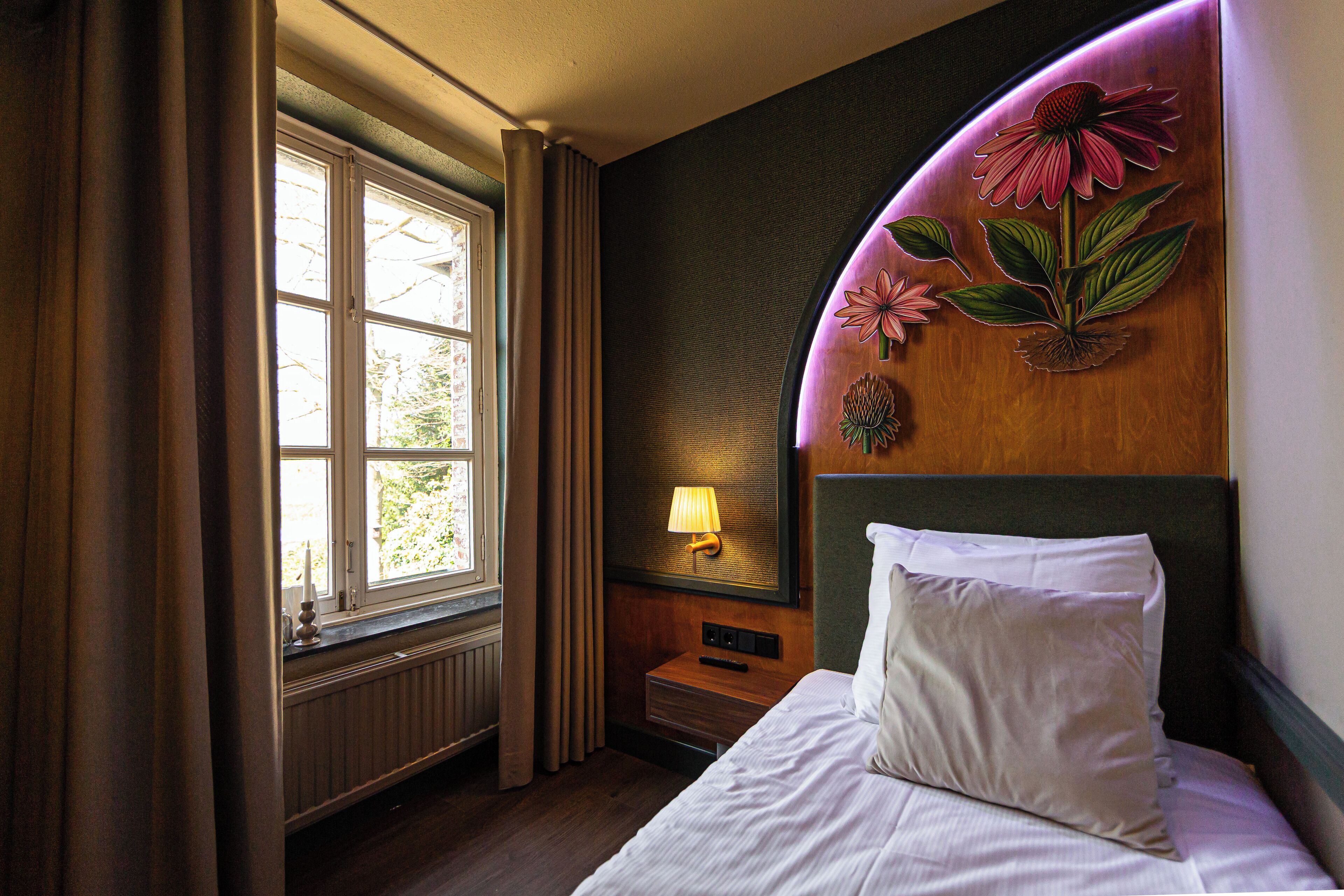 Quarto de hotel com cama, janela com cortinas, luminária fixada na parede e painel floral decorativo.