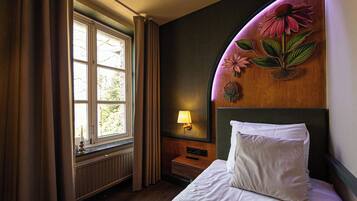 Quarto de hotel com cama, janela com cortinas, luminária fixada na parede e painel floral decorativo.