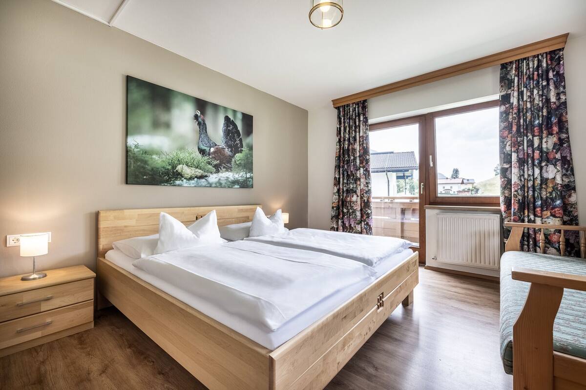 Quarto de hotel com cama de casal, mesa de cabeceira, luminária, janela grande com cortinas e vista exterior.