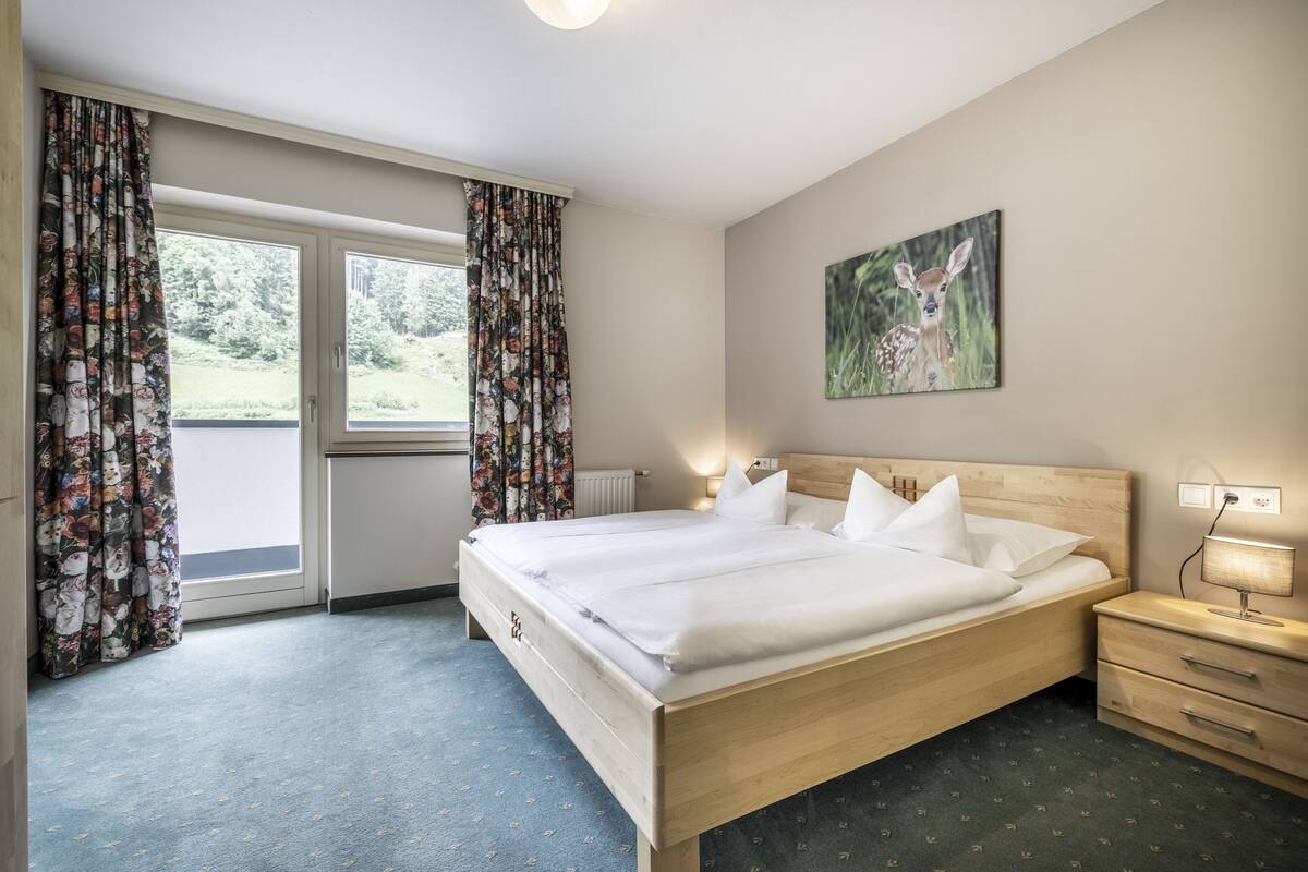 Quarto com cama de madeira, mesa de cabeceira, abajur, pintura de um veado e vista para o verde pela janela.