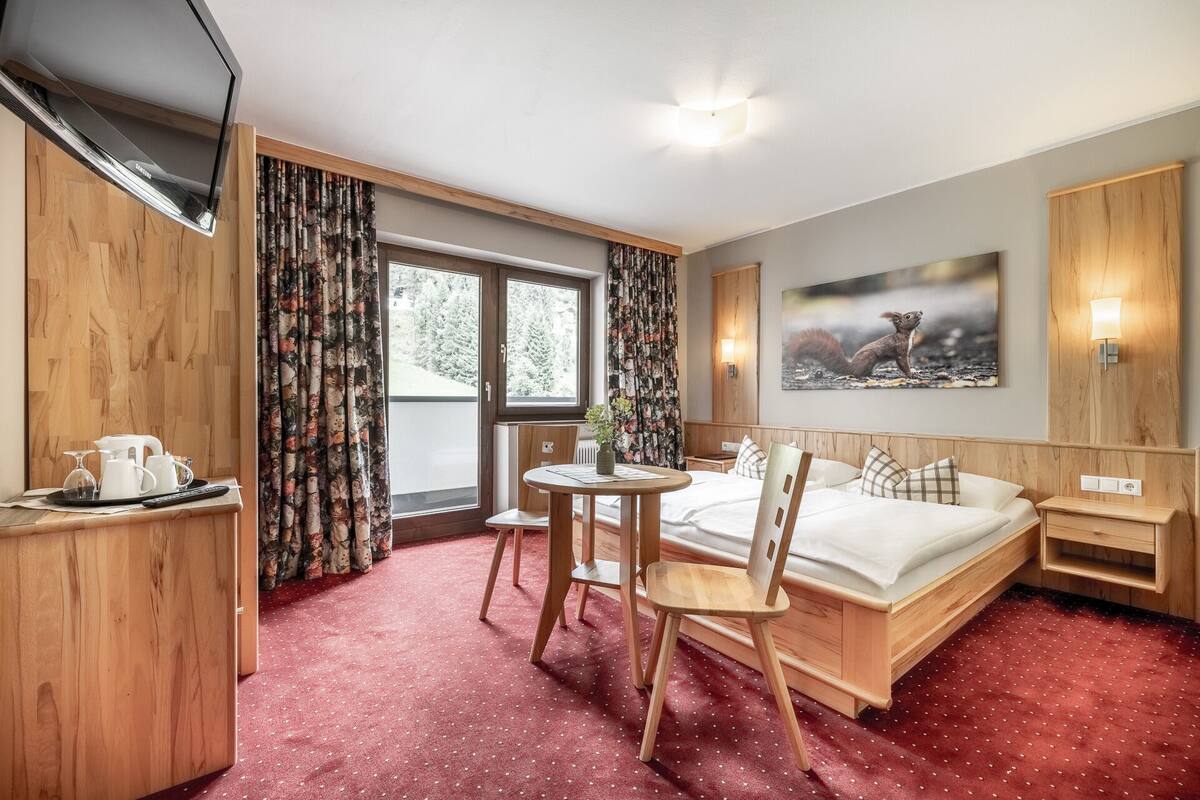 Quarto de hotel com cama, escrivaninha, cadeira, mesinha e janela com cortinas.