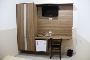 Quarto compacto com escrivaninha embutida, televisão de tela plana, mini-geladeira e lixeira.