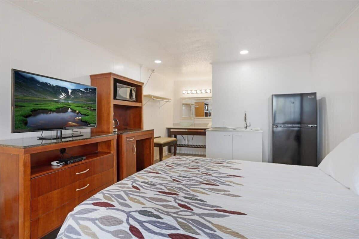 Quarto de hotel com cama, televisão, armário de madeira, frigorífico e uma pequena cozinha.