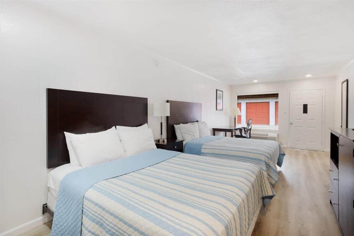 Quarto de hotel com duas camas, uma escrivaninha, uma cadeira e uma janela com persianas.