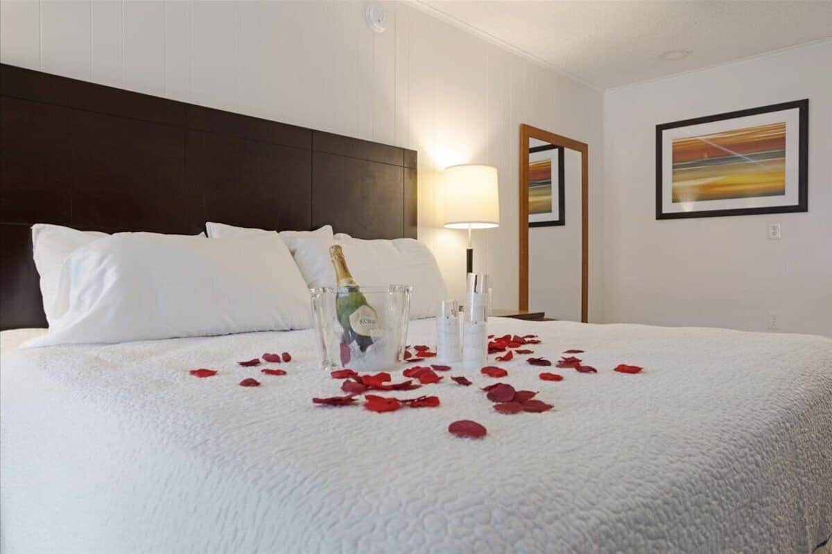 Quarto de hotel com uma cama grande, roupa de cama branca, uma garrafa de champanhe e pétalas de rosa na cama.