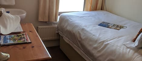 Quarto de hotel com cama, escrivaninha, cadeira, luminária, janela com cortinas e radiador.